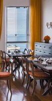 Uma sala de jantar com mesa posta para quatro, cadeiras de madeira, um armário com gavetas e vista para o mar através de uma grande janela.