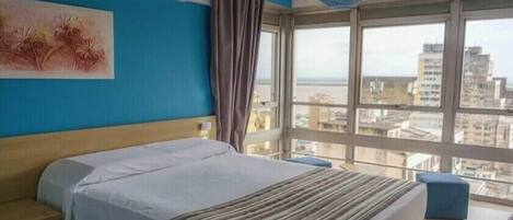 Quarto de hotel com uma cama grande, vista da cidade através de amplas janelas e uma parede azul.