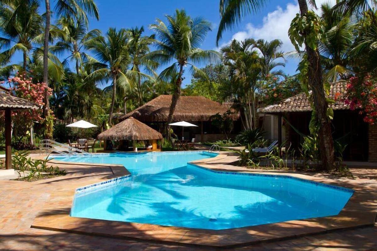 Uma área de piscina com várias piscinas, palmeiras e estruturas com telhados de palha.