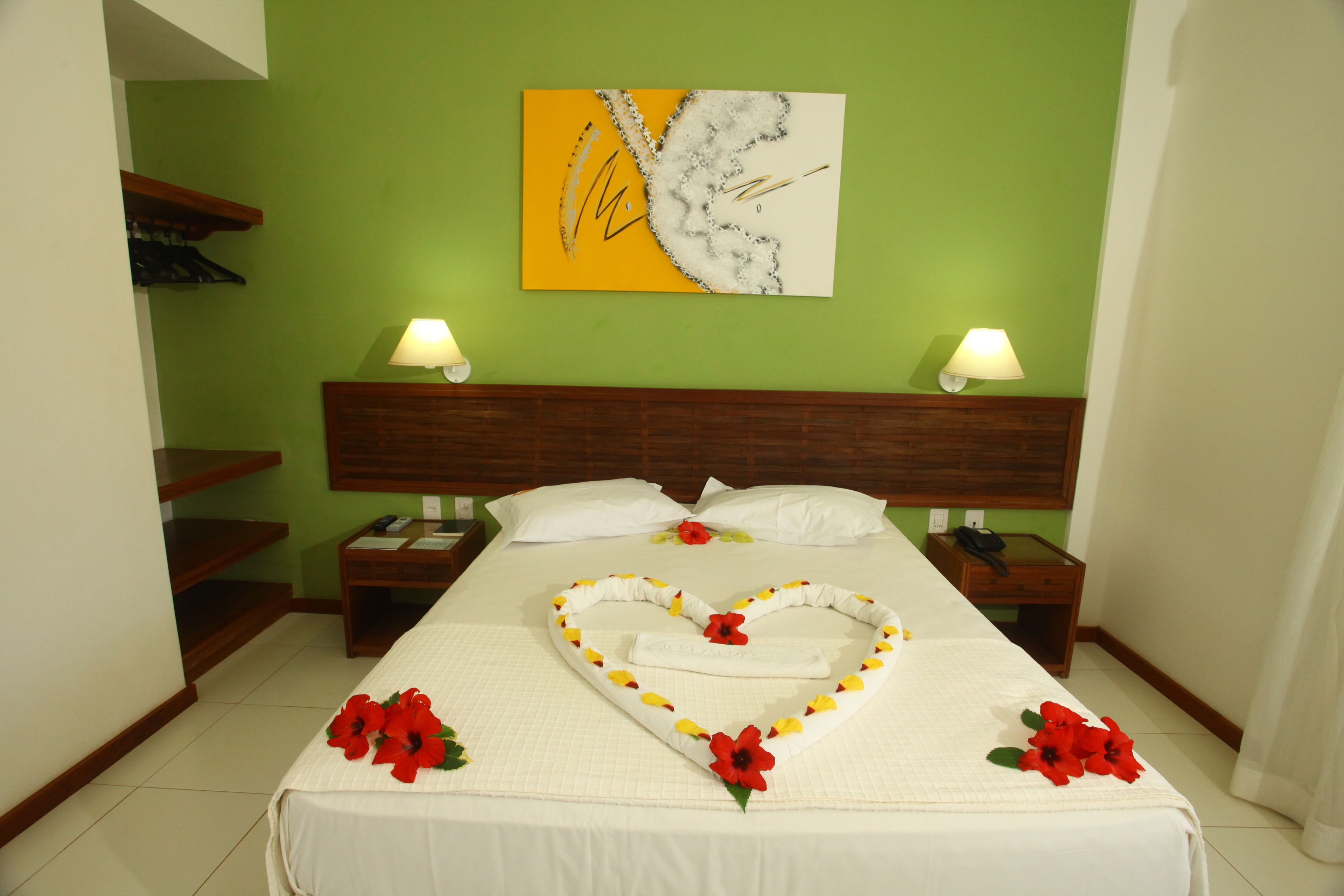 Quarto de hotel com uma cama, duas mesas de cabeceira, um criado-mudo, uma obra de arte na parede e uma decoração em formato de coração na cama.