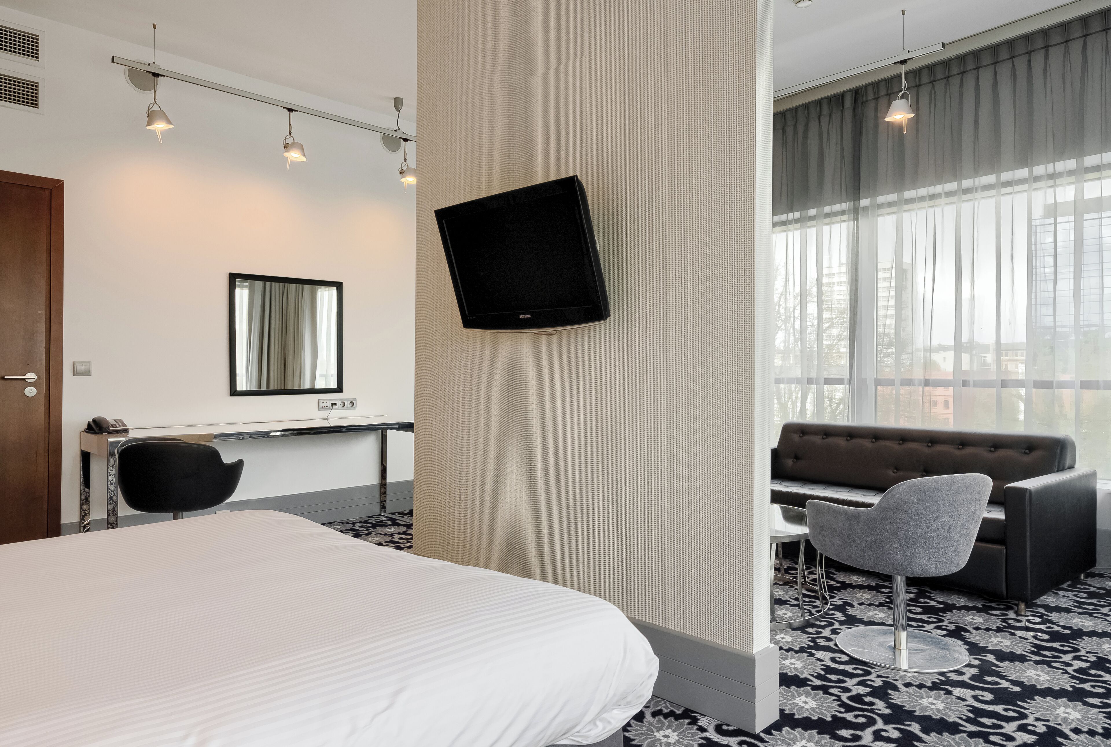 Quarto de hotel com uma cama grande, uma escrivaninha com cadeira, um sofá, televisão fixada na parede e uma janela com cortinas.