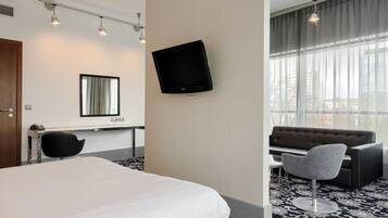 Quarto de hotel com uma cama grande, uma escrivaninha com cadeira, um sofá, televisão fixada na parede e uma janela com cortinas.