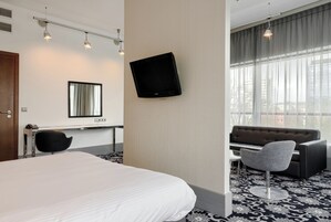 Quarto de hotel com uma cama grande, uma escrivaninha com cadeira, um sofá, televisão fixada na parede e uma janela com cortinas.