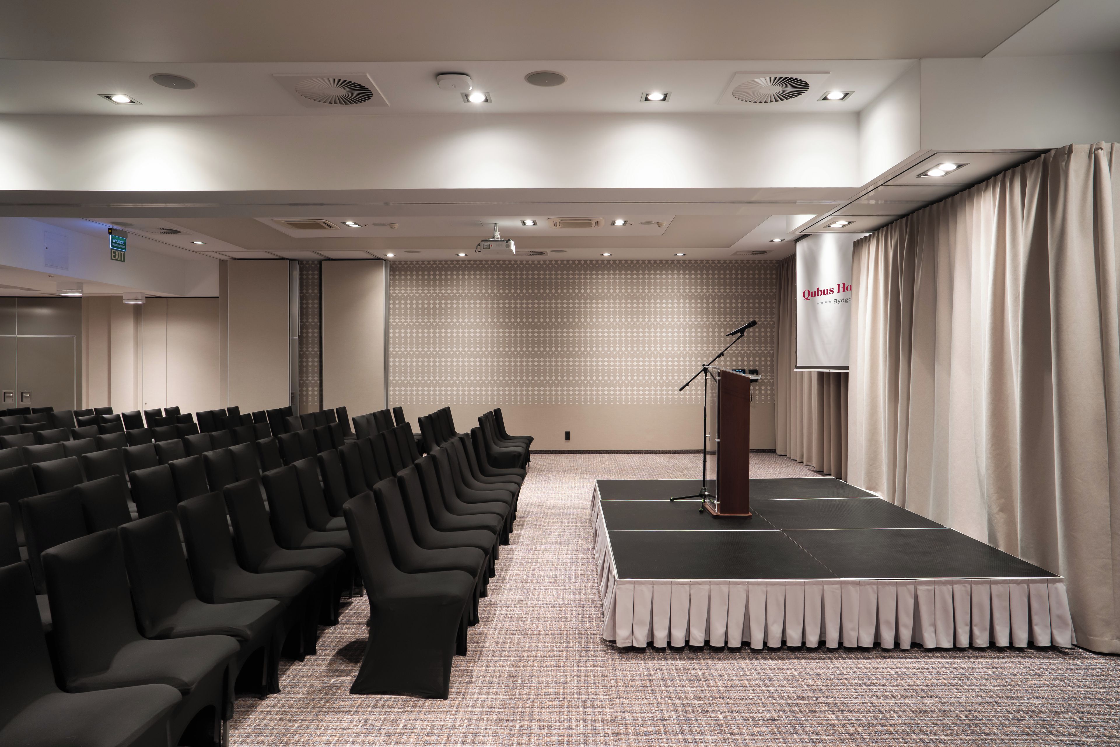 Uma sala de conferências com fileiras de cadeiras, um palco com um pódio e uma grande cortina.