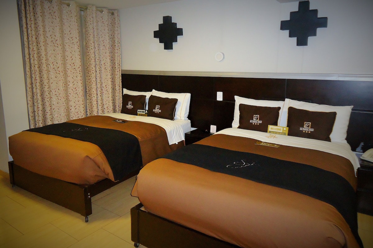 Quarto de hotel com duas camas, cada uma com espalha individual marrom e travesseiros brancos, e uma decoração na parede em formato de cruz preta.