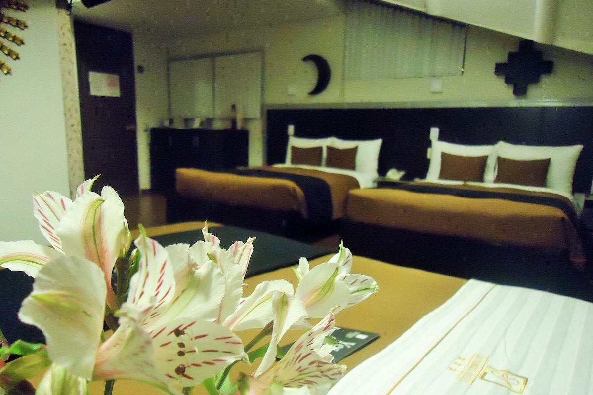 Quarto de hotel com duas camas, uma mesinha de cabeceira e um relógio na parede.