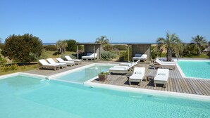 Área da piscina com espreguiçadeiras e deck de madeira, com vista para um céu azul límpido e oceano.
