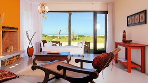 Uma sala de estar com sofá, poltrona, mesa de centro e lareira. Há vista para a praia pelas portas de vidro.