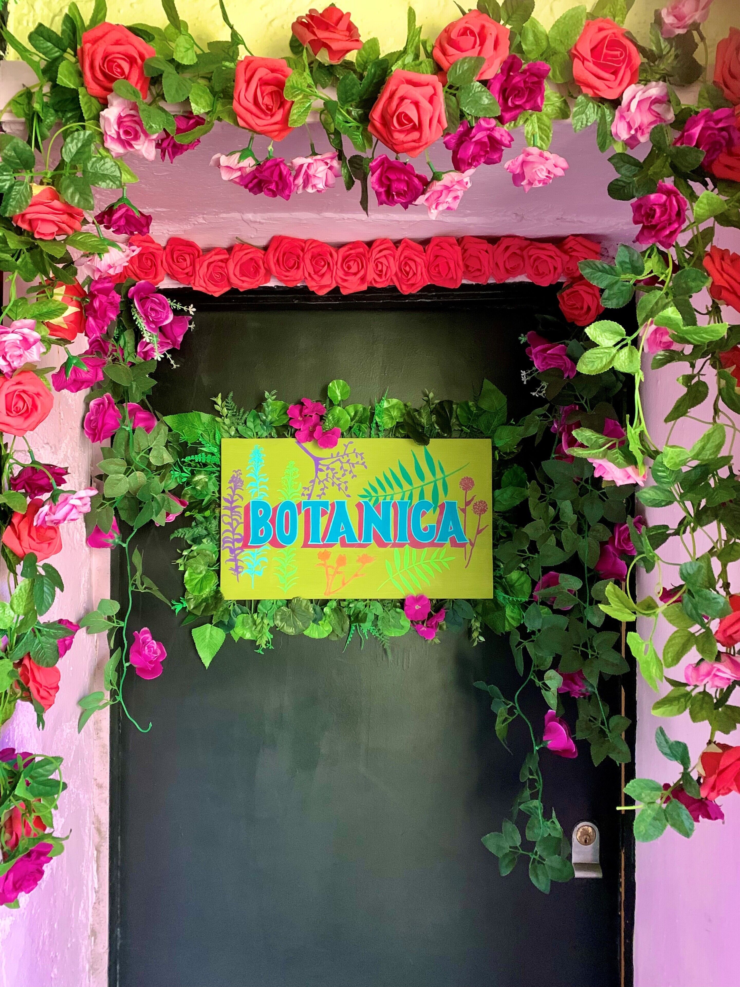 Uma porta com uma placa que diz "BOTANICA", rodeada por um arranjo floral de rosas e folhagem.