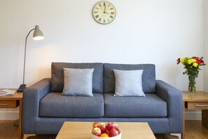 Uma sala de estar moderna com um sofá cinzento, duas almofadas brancas, uma mesa de centro de madeira com uma tigela de frutas, um relógio de parede, um abajur e um vaso com flores.