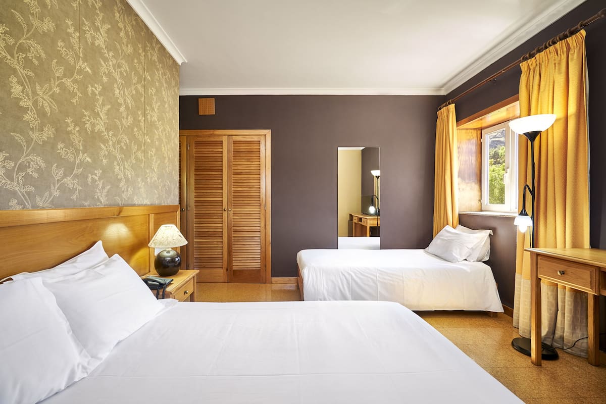Quarto de hotel com duas camas, um guarda-roupa de madeira, uma escrivaninha com abajur e uma janela com cortinas.