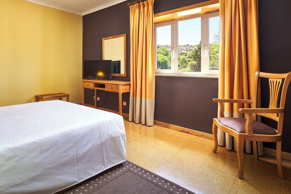 Quarto de hotel com cama, escrivaninha com computador, cadeira e janela com cortinas.