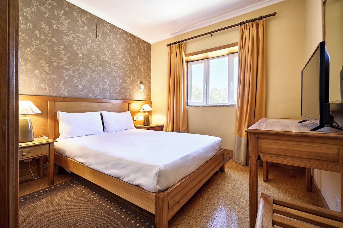 Quarto de hotel com cama de madeira, luminárias de cabeceira, escrivaninha e televisão.