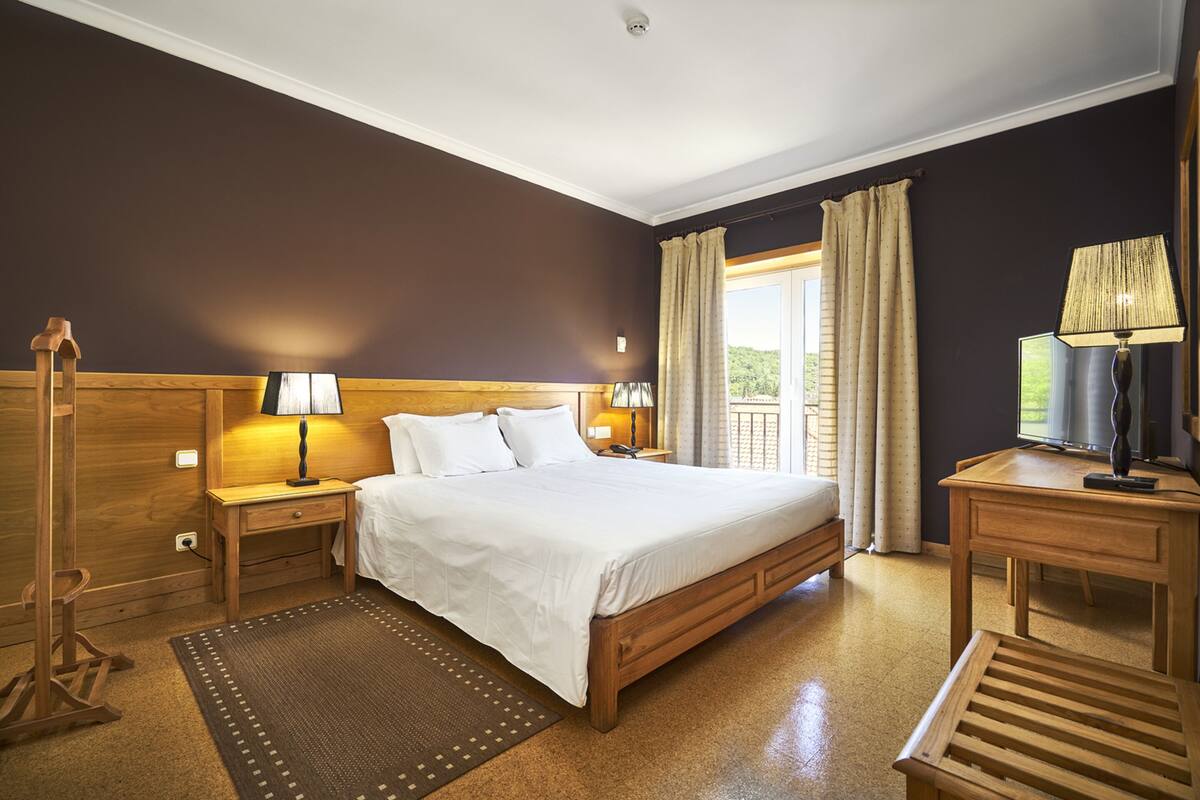 Quarto com cama grande, cabeceira de madeira, mesas de cabeceira com abajures, um banco de madeira e vista para o exterior através de uma janela.
