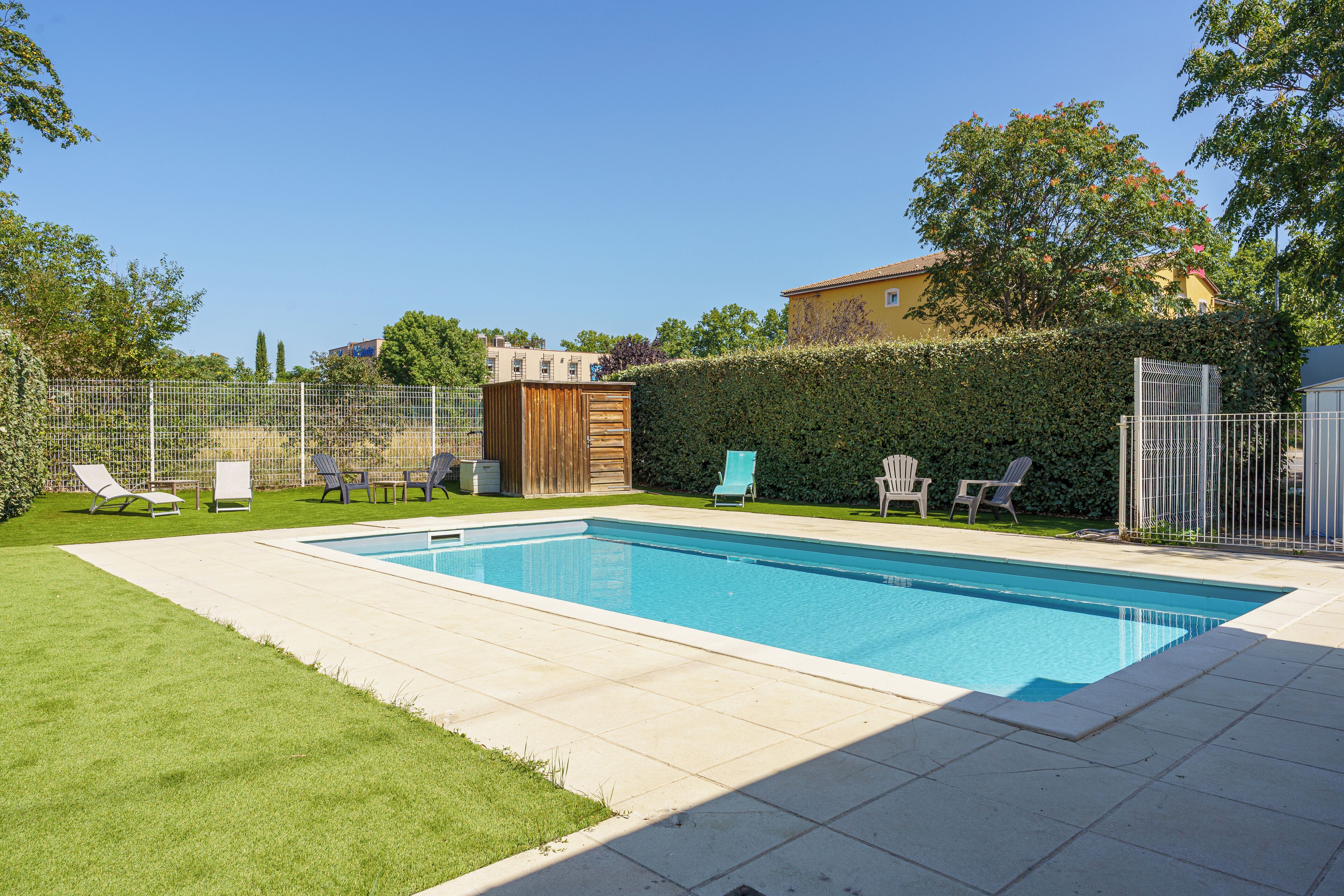 Piscina retangular com deck ladrilhado, rodeada por um sebe baixo e uma cerca de metal.
