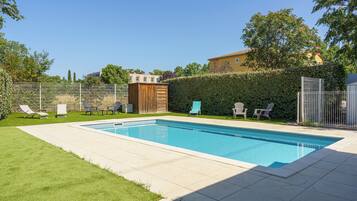 Piscina retangular com deck ladrilhado, rodeada por um sebe baixo e uma cerca de metal.