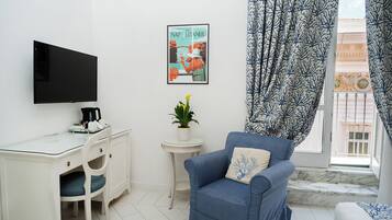 Quarto com televisão de tela plana, escrivaninha com gavetas, cadeira, pequena mesa com uma planta e sofá azul com almofada.
