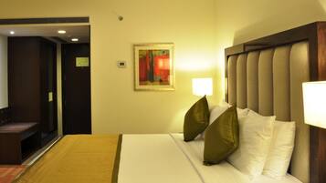 Quarto de hotel com uma cama grande, cabeceira com ripas verticais, um quadro na parede e uma porta que dá para outro quarto.