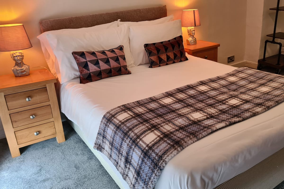 Cama bem arrumada com manta xadrez, duas almofadas e duas luminárias de cabeceira. Um criado-mudo de madeira com gavetas e uma pequena estátua.