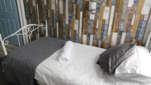 Uma cama de solteiro com colchão branco, manta cinza e uma toalha branca enrolada. Um travesseiro cinza de tricô está sobre a cama. A parede ao fundo apresenta um padrão de ripas de madeira.