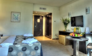 Quarto de hotel com uma cama, uma escrivaninha com uma cesta de frutas, uma televisão e uma porta que dá para outro quarto.