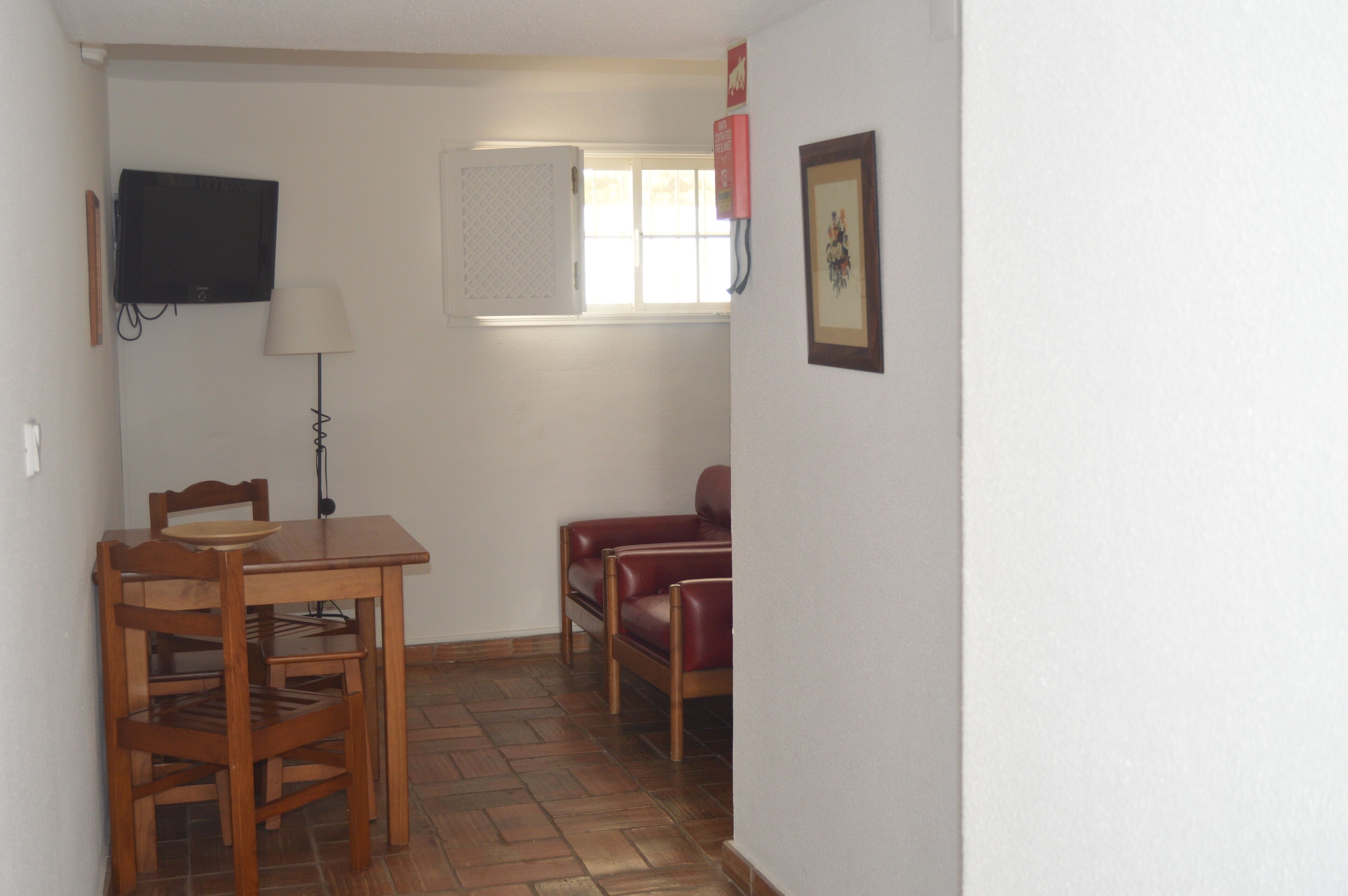 Um quarto pequeno com uma mesa de madeira, duas cadeiras, uma televisão, uma janela com persianas e um quadro na parede.