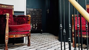 Uma escadaria com corrimão preto e balaustrada de madeira, que leva a um quarto escuro com uma poltrona estofada vermelha e um tapete estampado.