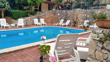 Piscina com cadeiras de descanso brancas e parede de pedra.