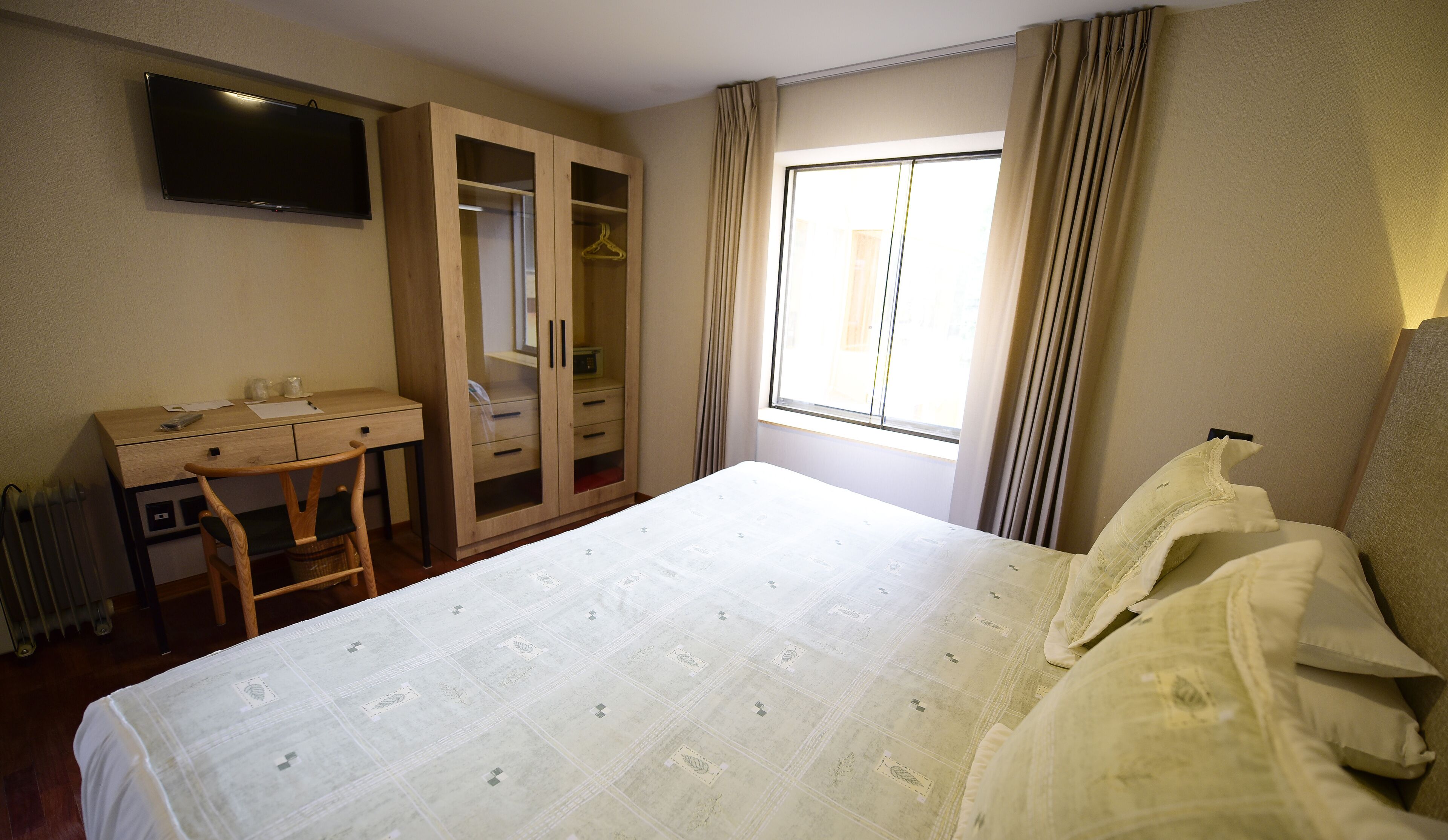 Quarto de hotel com cama, escrivaninha com cadeira, guarda-roupa, televisão e janela com cortinas.