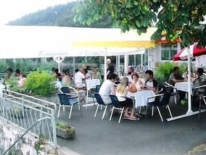 Área de refeições ao ar livre com mesas e cadeiras, cercada por vegetação e um edifício ao fundo.