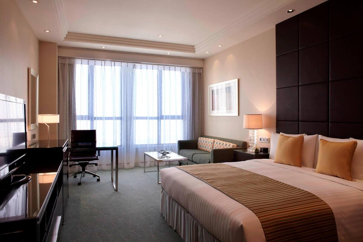 Quarto de hotel com uma cama grande, uma escrivaninha, uma cadeira, uma televisão e uma janela com cortinas.