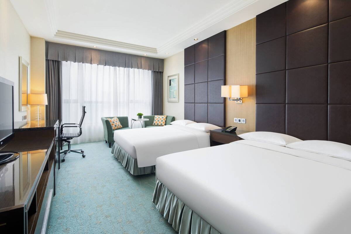 Quarto de hotel com duas camas, uma escrivaninha, uma cadeira, televisão e uma janela grande com cortinas.