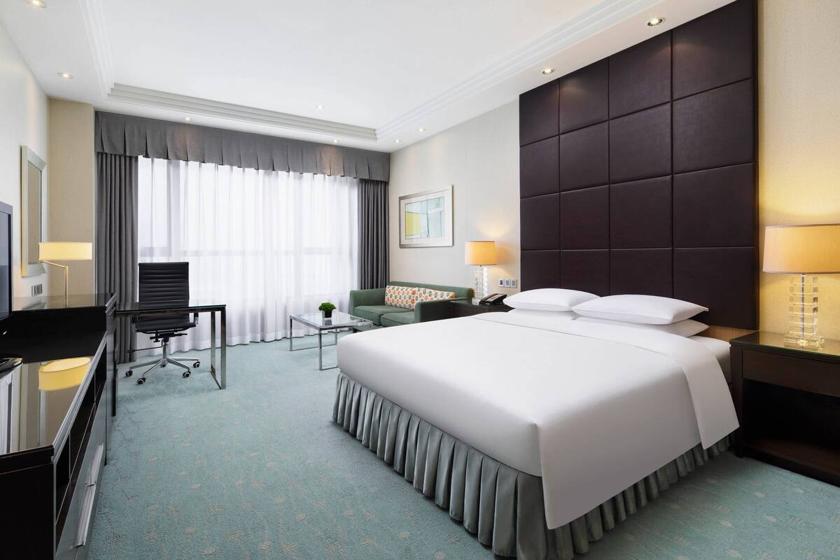 Quarto de hotel com uma cama grande, uma escrivaninha com cadeira, um sofá, uma televisão e uma janela com cortinas.