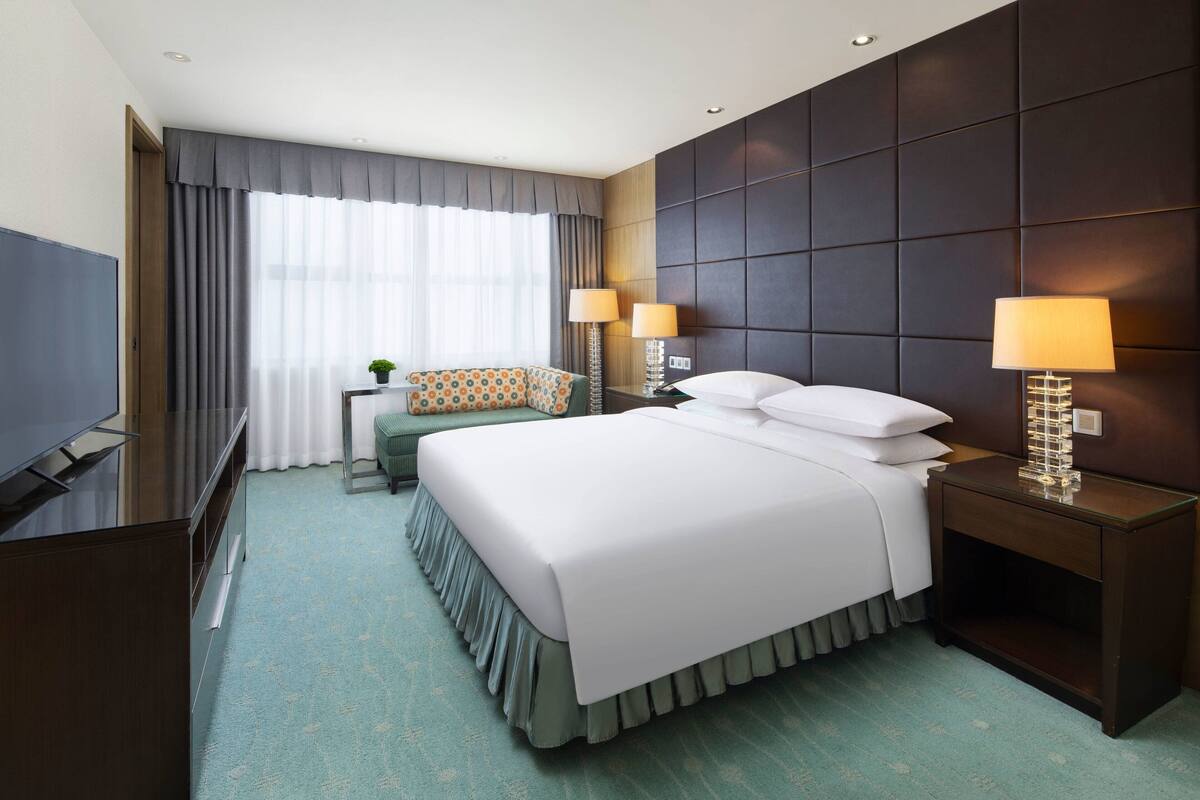 Quarto de hotel com uma cama grande, duas mesas de cabeceira com abajures, uma mesinha com uma planta e uma televisão em um suporte.
