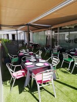 Área de refeições ao ar livre com mesas e cadeiras, protegida por um toldo.