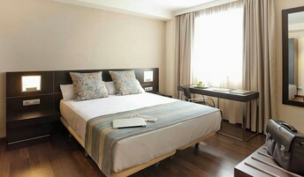Quarto de hotel com cama, mesas de cabeceira, escrivaninha, cadeira, mala e uma janela grande com cortinas.