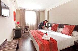 Quarto de hotel com cama, escrivaninha, cadeira, televisão e janela com cortinas.