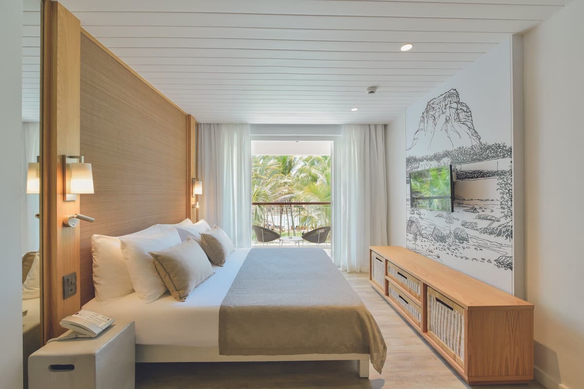 Quarto de hotel moderno com uma cama grande, cabeceira de madeira, mesa de cabeceira com abajur e vista para o exterior através de uma varanda.