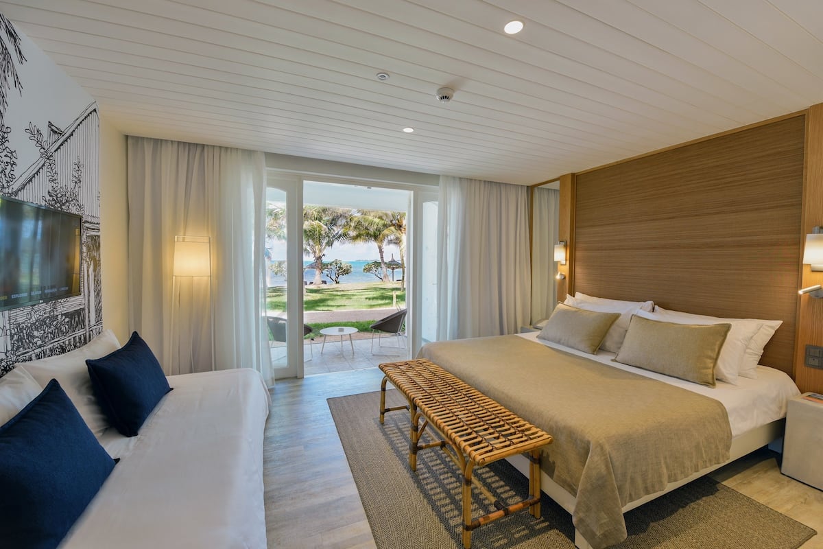 Quarto de hotel com cama grande, sofá, banco de madeira, TV e vista para o exterior.