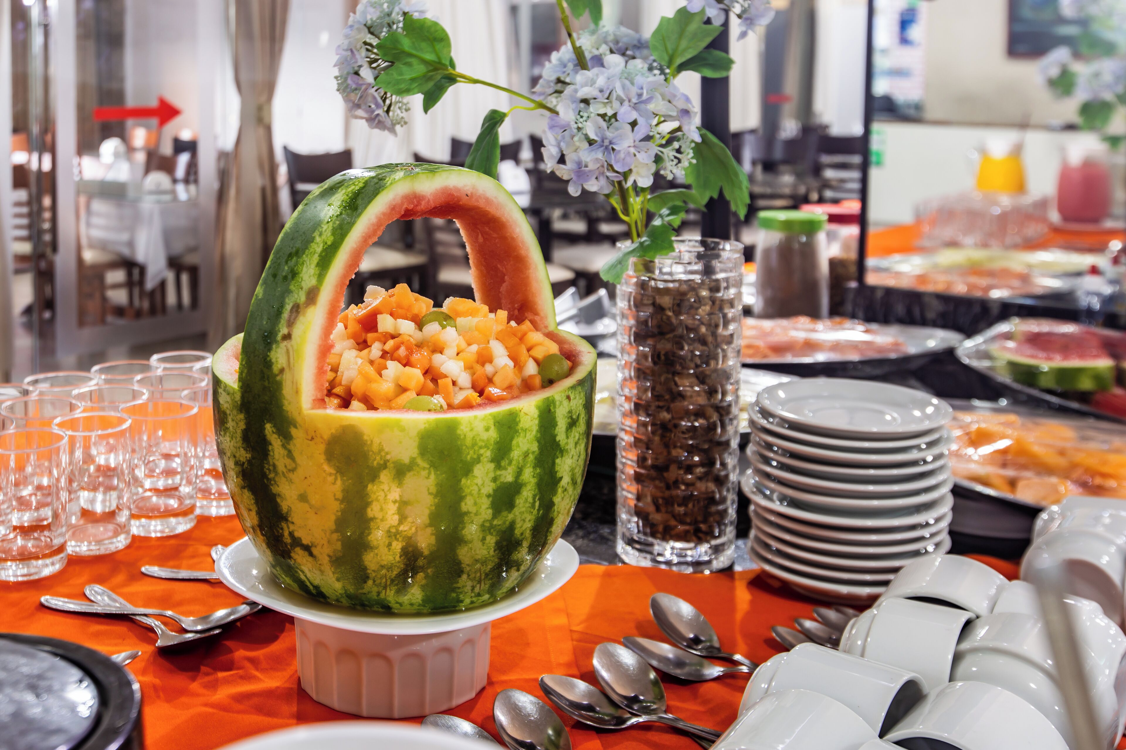 Mesa de buffet com uma grande tigela de melancia cheia de frutas picadas, cercada por pratos, recipientes e talheres.