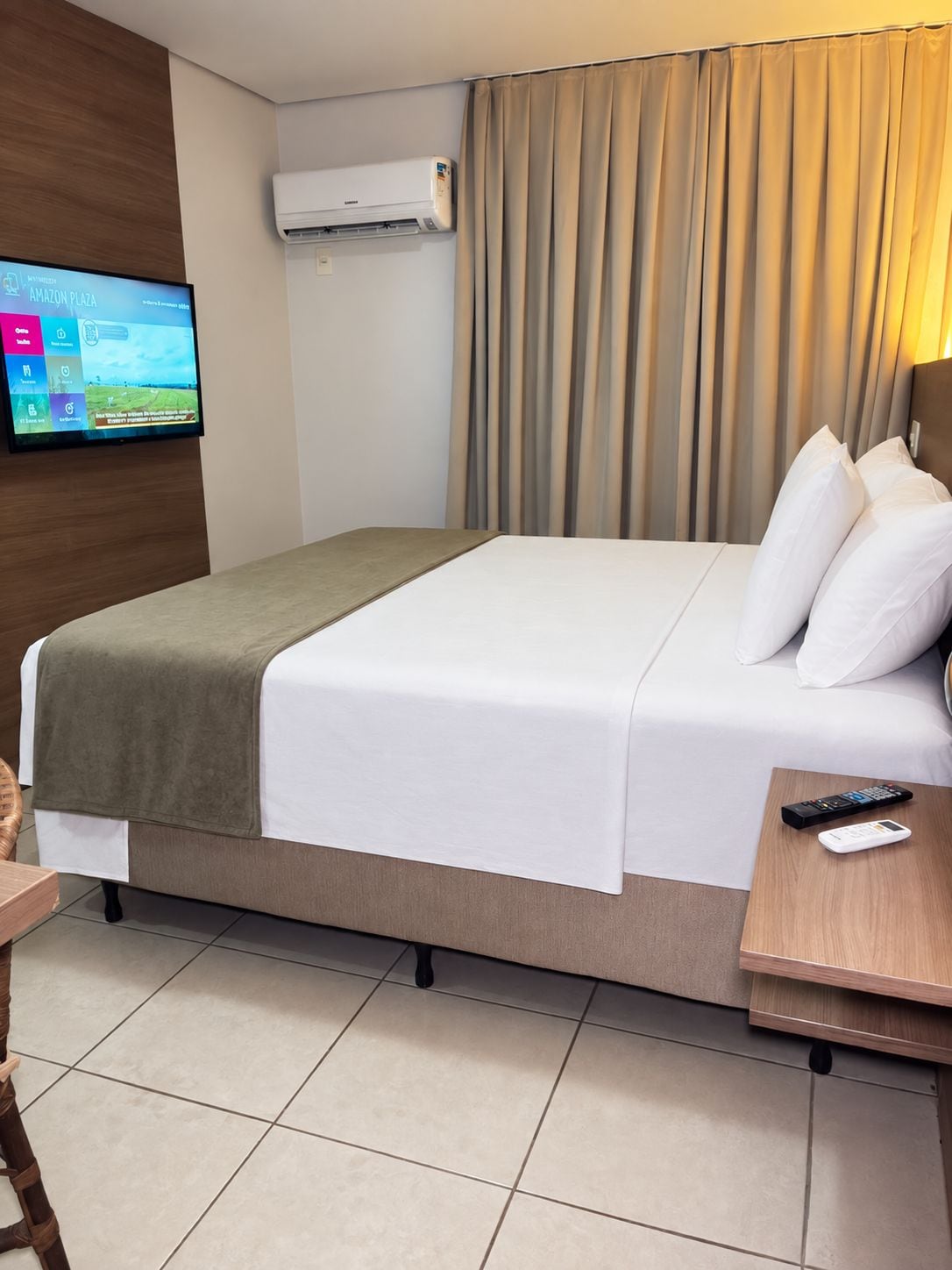 Quarto de hotel com uma cama, televisão na parede, controle remoto e abajur em uma mesa de cabeceira.