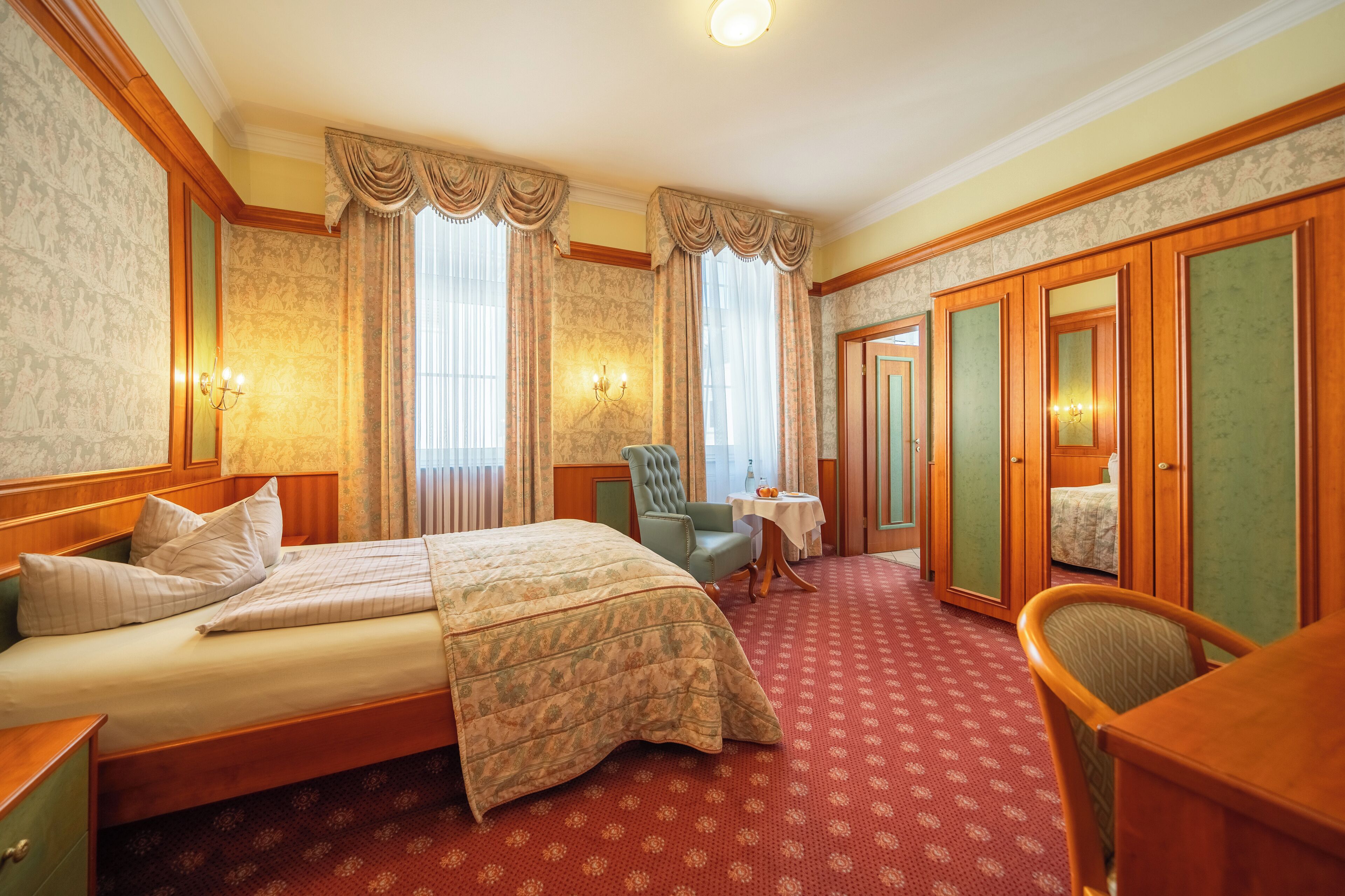 Quarto de hotel com cama, uma cadeira, uma mesinha e um guarda-roupa.
