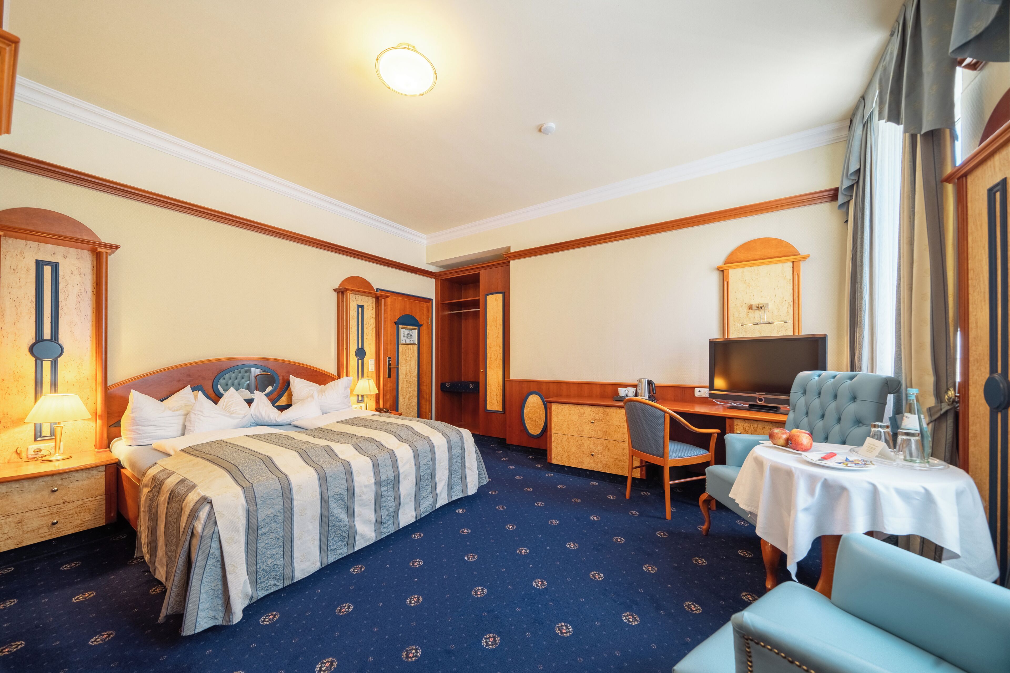 Quarto de hotel com uma cama grande, uma escrivaninha com computador, uma cadeira, uma mesa pequena com abajur e uma televisão.