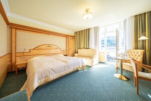 Quarto de hotel com uma cama grande, um espaço de estar com uma cadeira e mesa, e vista para a cidade pela janela.