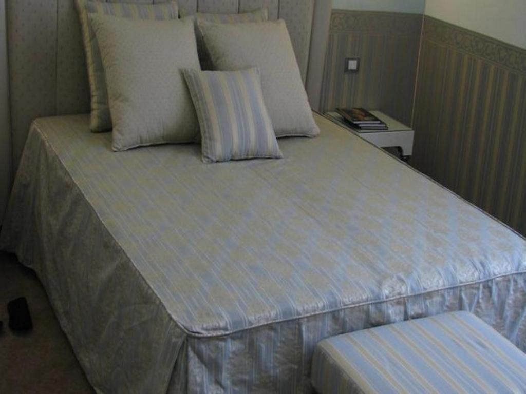 Uma cama com colcha listrada e almofadas, uma mesa de cabeceira com um livro, e um criado-mudo com uma luminária.