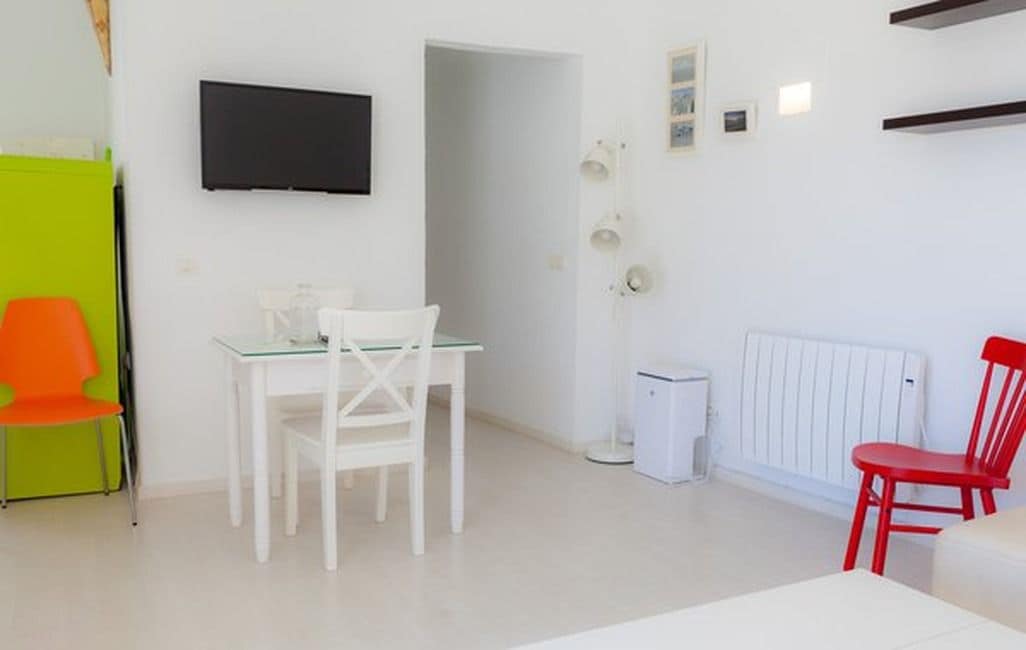 Quarto com uma mesa e cadeiras brancas, televisão fixada na parede, radiador e porta que dá para outro cômodo.