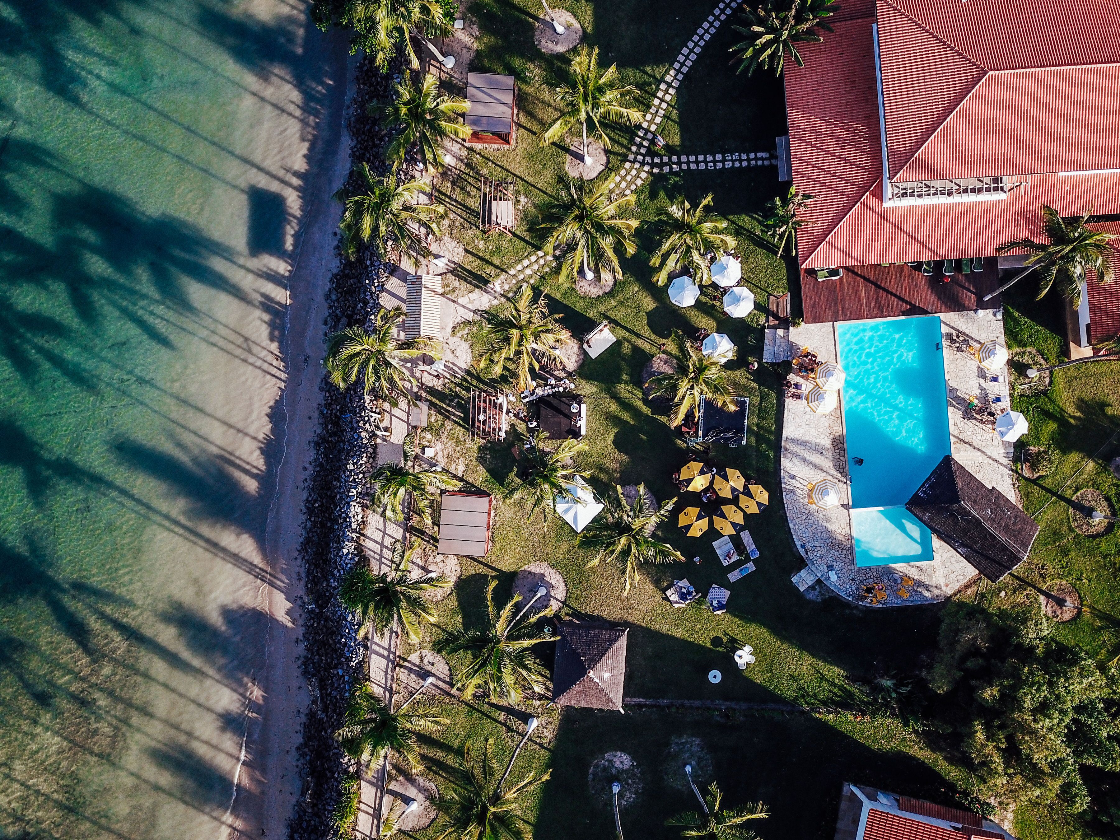 Vista aérea de um resort com piscina, palmeiras e assentos à beira-mar.