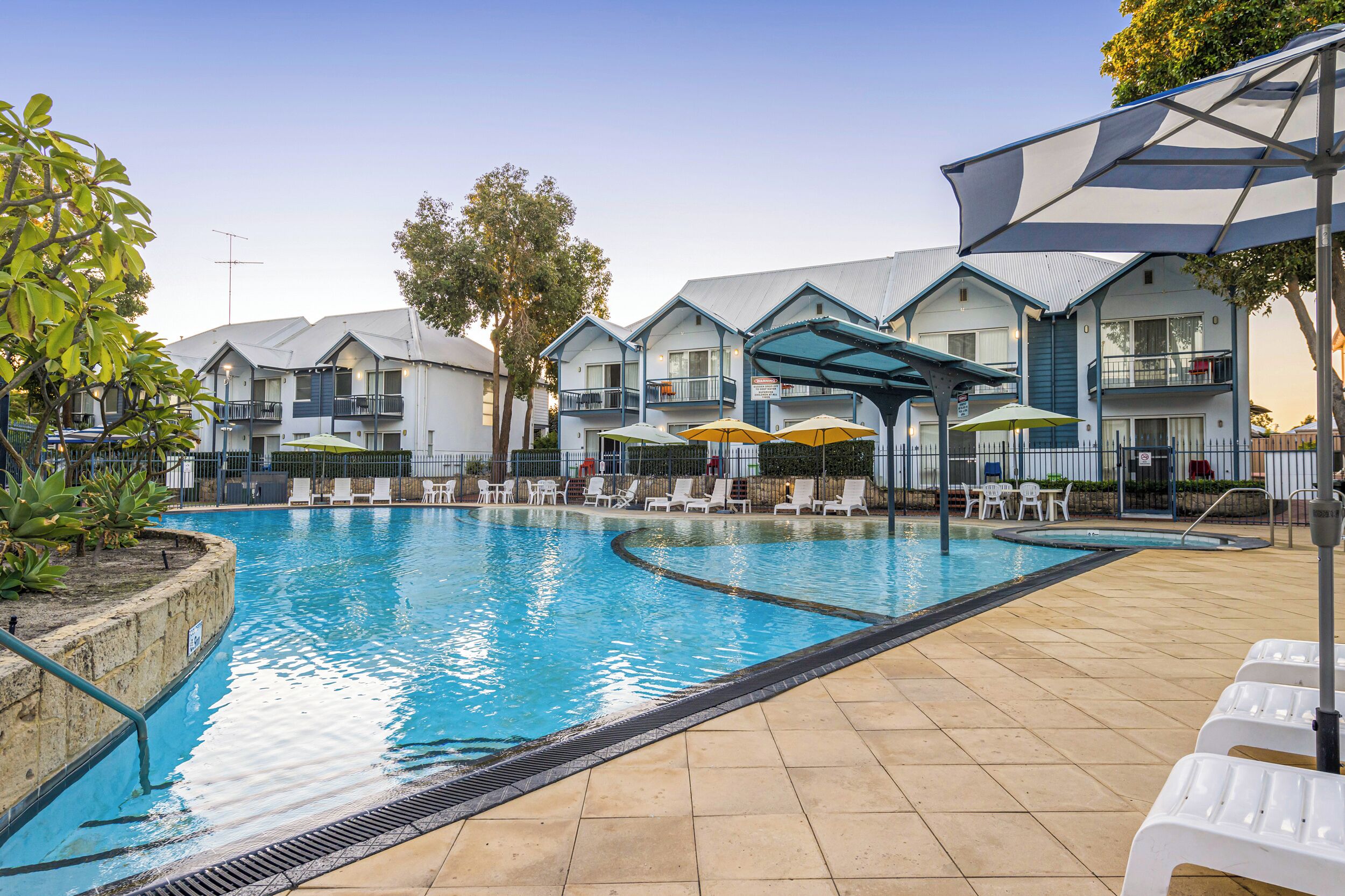 Piscina com espreguiçadeiras e guarda-sóis, cercada por edifícios com varandas e céu claro.