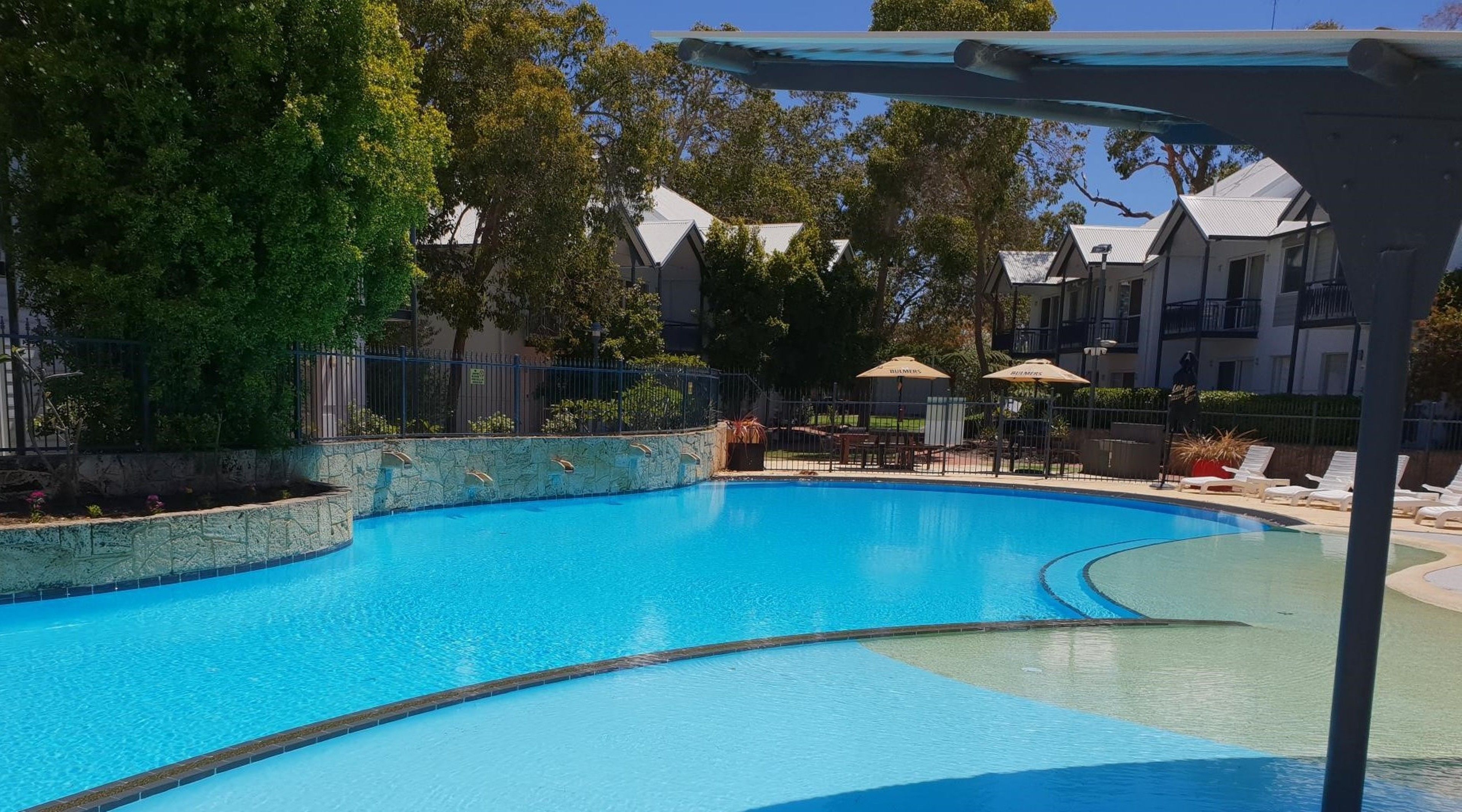 Piscina com borda de pedra, rodeada por espreguiçadeiras e guarda-sóis, com edifícios residenciais ao fundo.