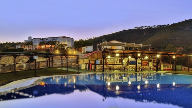 Piscina com área de bar e assentos externos, rodeada por edifícios e colinas.